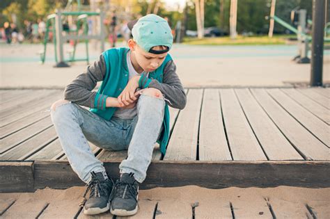 어린 시절 무릎 부상 스포츠 경기장에서 무릎을 꿇고 모자를 쓴 십대 청소년이 청바지를 입고 무릎을 다쳤습니다 그는 무릎을 바라본다 복사 공간 긁힌에 대한 스톡 사진 및 기타