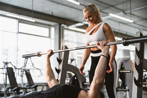 젊은 남성 운동 선수는 여성 코치와 스포츠 클럽에 종사하고 있습니다 한 남자가 가슴에서 벤치 프레스를 수행합니다 강사는 그 사람을 보장합니다 여자에 대한 스톡 사진 및 기타