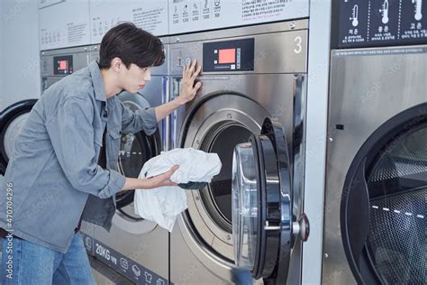 Foto Stock 셀프 서비스 세탁실에서 세탁기 또는 건조기 앞에서 세탁물을 들고 냄새를 맡거나 고민하고 있는 젊은 아시아