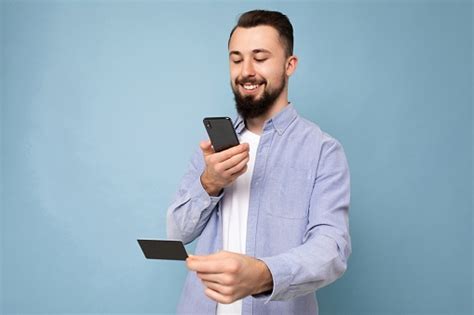 검은 신용 카드를 들고 휴대 전화로 사진을 만들기위한 복사 공간이있는 파란색 배경 벽에 고립 된 캐주얼 세련된 파란색 셔츠와