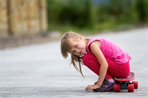 캐주얼 핑크 의류꽤 젊은 긴 머리 금발의 아이 소녀는 흐린 여름 녹색 배경에 햇볕이 잘 드는 거리에 스케이트 보드에 손에 기대어 앉아있다 어린이 활동 게임 및 재미있는 개념
