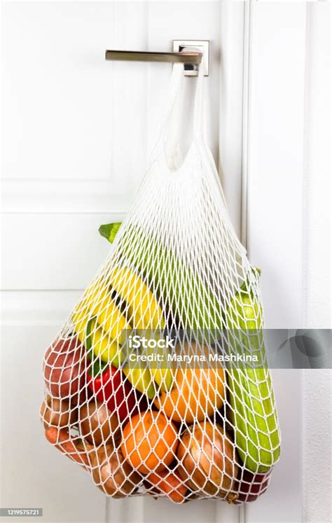 과일과 채소는 끈 가방에 흰색 문 손잡이에 매달려 있습니다 양파 호박 바나나 후추 오렌지 에코 제로 폐기물 개념 플라스틱이