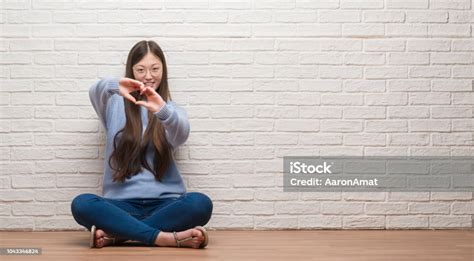 젊은 중국 여자 마음 기호와 손으로 모양을 보여주는 사랑에 웃 고 벽돌 벽을 통해 바닥에 앉아 낭만적인 개념입니다 개념에 대한 스톡 사진 및 기타 이미지 Istock