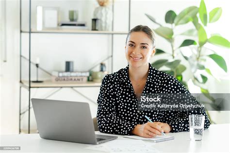 행복한 성공적인 사업가 관리자 또는 비서는 노트북을 들고 책상에 앉아 펜을 들고 카메라를 바라 보며 현대적인 사무실에서 자신감