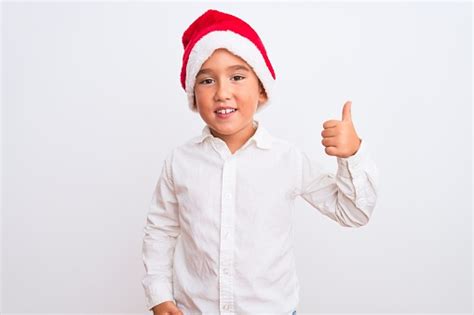 고립 된 흰색 배경 위에 서있는 크리스마스 산타 모자를 쓰고있는 아름다운 아이 소년은 행복한 엄지 손가락으로 손을 들고 제스처를 취합니다 성공을 보여주는 카메라를 보고
