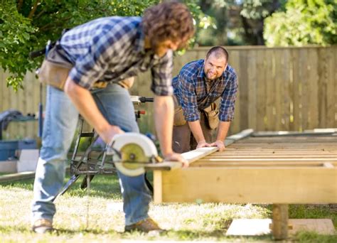 Carpenter Cutting Wood With Saw While Coworker Drilling At Site Stock