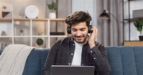 온라인 수업 E 러닝 개념 젊은 호감 가는 인도 남자는 집 거실의 소파에 앉아 노트북과 헤드셋을 사용하여 공부하고 화면을