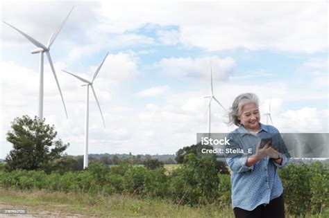 풍력 터빈 농장에서 스마트폰을 들고 있는 나이 든 아시아 여성 풍차 공원에서 휴대 전화를 사용하는 노인 여성 기술 생태학 대체 재생 에너지 및 환경으로 능동적으로 성숙합니다