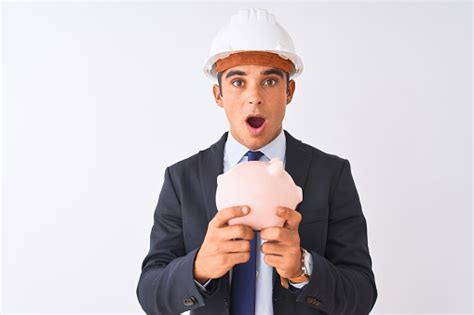 고립 된 흰색 배경 위에 돼지 저금통을 들고 헬멧을 쓰고 있는 잘생긴 건축가 남자는 깜짝 얼굴로 충격에 무서워하고 두려움과 두려움 표현으로 흥분했습니다 유머에 대한 스톡