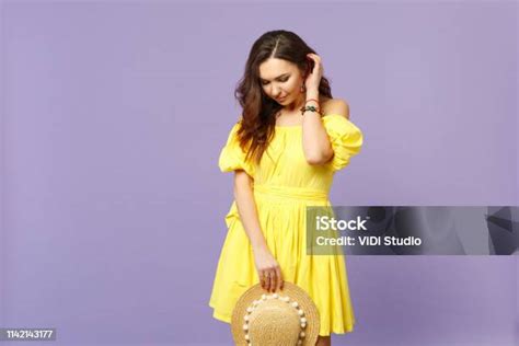 여름 모자를 들고 노란 드레스에 예쁜 젊은 여자 스튜디오에서 파스텔 바이올렛 배경에 고립 된 머리에 손을 유지 하 고 찾고 사람들은 진심으로 감정 라이프 스타일 개념입니다