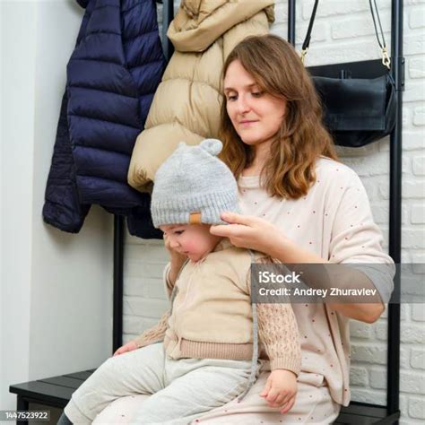 어머니는 집 복도에 앉아있는 유아 아기의 머리에 따뜻한 모자를 씌 웁니다 여자 엄마는 추운 날씨에 겨울 산책을 위해 아이에게 따뜻한 옷을 입는다 1세 3개월 된 아이 가을에