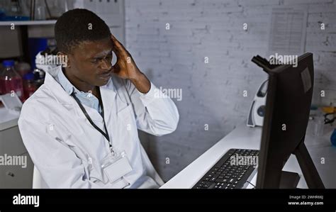 Stressed African Man In Lab Coat Analyzing Data On Computer In Modern Laboratory Room Stock