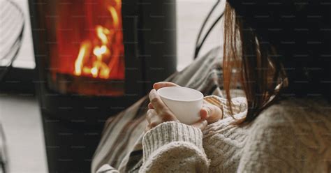 따뜻한 차 한 잔을 들고 산의 전망을 감상할 수 있는 현대적인 검은 벽난로에서 휴식을 취하는 세련된 여성 추운 계절에 아늑하고 따뜻한 순간 니트 스웨터와 모자를 쓴 젊은