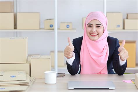 파란색 무슬림 정장을 입은 활동적인 아시아 여성이 앉아서 컴퓨터와 온라인 소포 상자 배달을 하고 있다 스타트업 중소기업 프리랜서 일하는 소녀가 흥미진진한 감정으로 엄지