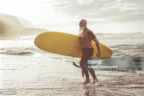 일몰에서 서핑 후 롱 보드와 함께 걷는 시니어 핏 남자 성숙한 문신 남자는 열대 해변에서 익스트림 스포츠를하는 재미를 즐거운 노인 라이프 스타일과 여행 개념 남성의 몸에