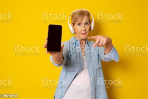 노란 배경위에 고립된 모바일 화면을 보여주는 헤드폰을 착용한 성숙한 여성 가리키기에 대한 스톡 사진 및 기타 이미지 가리키기 공란 광고 Istock