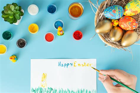 부활절을 위해 준비 하 고 손으로 만든 바구니는 여러 가지 빛깔의 페인트 옆에 부활절 달걀을 만들고 파란 배경이에 흰 종이에 행복 한 부활절을 칠하는 브러시를 사용 합니다