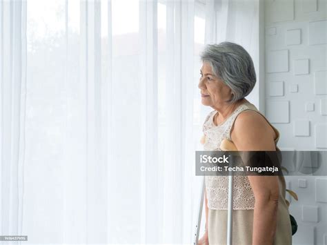 아시아 노인 여성 흰 머리카락은 커튼 근처에서 목발을 걷고 서서 복사 공간이있는 유리 창을 바라 봅니다 목발 보행기를 사용하는 노인 여성 환자 강력한 건강 의료 개념 65