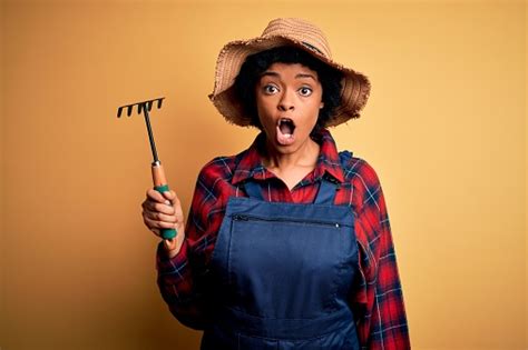 젊은 아프리카계 미국인 Afro 농부 여자 곱슬 머리를 입고 앞치마와 모자를 사용 하 여 레이크를 사용 하 여 충격에 무서워 하는 깜짝 얼굴 두려워 하 고 두려움 표정으로