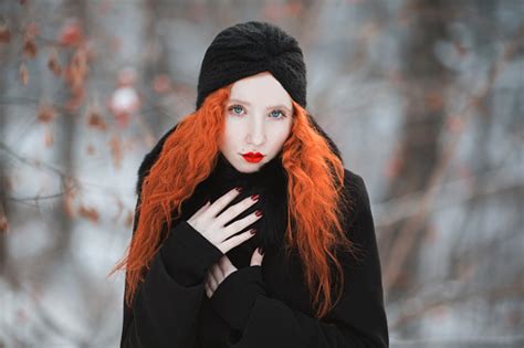 겨울 숲의 배경에서 모피와 함께 검은 코트에 붉은 머리를 가진 여자 창백한 피부와 그녀의 머리에 터 번을 가진 밝은 특이 한 외관으로 파란 눈 빨간 머리 소녀 여자의 스타일