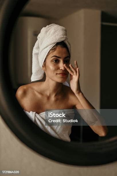 아름다운 젊은 여성은 아침 스킨케어 루틴으로 하루를 시작하고 욕실 거울을 바라보며 빛나는 얼굴에 크림을 부드럽게 바르고 있습니다 25 29세에 대한 스톡 사진 및 기타