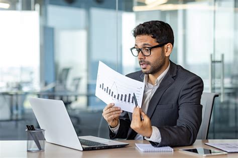 온라인 화상 회의 프레젠테이션에서 사무실 내부의 진지한 성공한 사업가 비즈니스 정장을 입은 남자가 카메라에 보고서 그래프를 보여주고 직장에서 화상 통화를 위해 노트북을