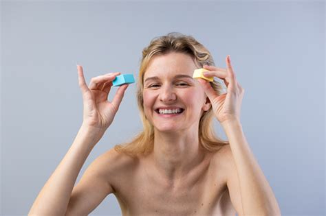 부드러운 회색 배경을 배경으로 양손에 형형색색의 지우개를 장난스럽게 들고 있는 쾌활한 젊은 여성은 얼굴에 밝은 미소를 지으며 기쁨과 긍정을 발산합니다 금발 머리에 대한 스톡