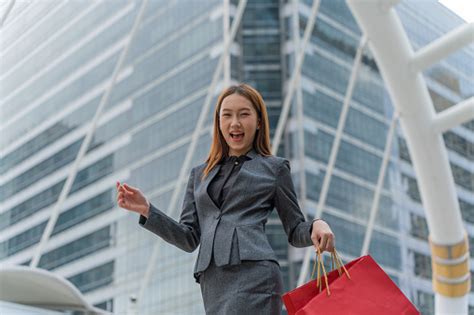 회색 사무실 정장을 입은 젊은 아시아 사업가가 빨간 쇼핑 종이 봉지를 손에 들고 중앙 비즈니스 지구를 행복하게 걷고 있다 18