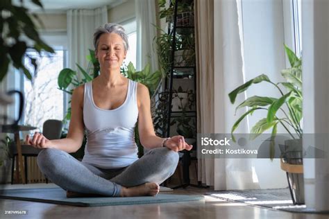 아름 다운 미소 백발의 성숙한 중년 여성이 집에서 연꽃 자세로 요가 매트에 앉아 있습니다 노인에 대한 스톡 사진 및 기타 이미지