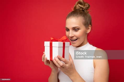 화려한 배경 벽에 고립 된 꽤 긍정적 인 놀란 젊은 금발 여성의 사진 사진은 선물 상자를 들고 선물 상자를 보고 빨간색 리본으로 선물 상자를보고 화려한 배경 벽을 통해 고립