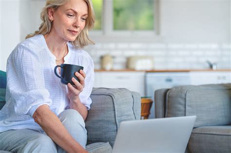 노트북 컴퓨터를 사용하고 소파에 앉아 차나 커피 한 잔을 즐기는 성숙한 여성 50 59세에 대한 스톡 사진 및 기타 이미지 50 59세 Brand Name Online