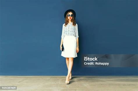 흰색 줄무늬 셔츠 검은 둥근 모자 진한 파란색 배경에 핸드백 클러치를 착용 한 전체 길이 아름다운 젊은 여성 20 29세에 대한
