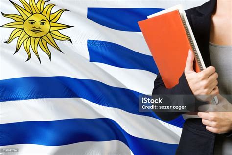 우루과이 언어 개념 학습 백그라운드에서 우루과이 국기와 함께 서 있는 젊은 여자 책을 들고 교사 오렌지 빈 책 표지 교사 교육 관련 직업에 대한 스톡 사진 및 기타 이미지