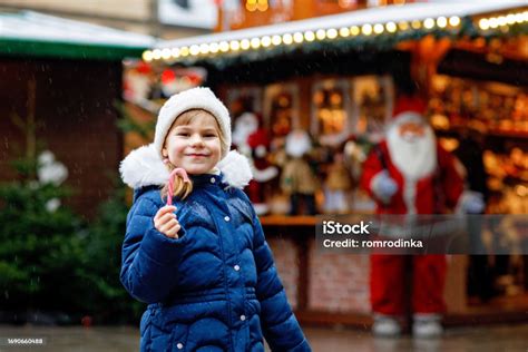 크리스마스 시장에 과자 가판대에서 사탕 지팡이를 가진 귀여운 미취학 소녀 독일의 전통적인 가족 시장에서 행복한 아이 눈이 내리는 동안 화려한 겨울 옷을 입은 미취학 아동