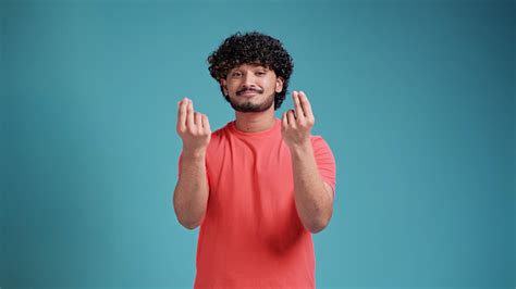 파란색 스튜디오 배경에 산호 티셔츠를 입고 수염을 기른 젊은 잘생긴 인도 남자가 손으로 돈 제스처를 하고 급여 지급을 요구한다 가리키기에 대한 스톡 사진 및 기타 이미지