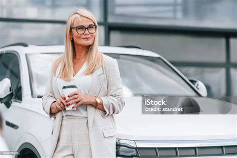 음료 한 잔과 함께 야외에서 흰색 정장을 입은 여자가 자동차 대리점에 있다 50 54세에 대한 스톡 사진 및 기타 이미지 50 54세 건물 외관 경영자 Istock