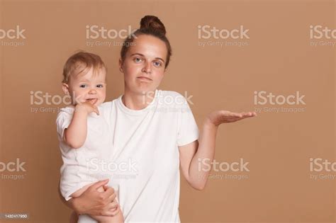 흰 티셔츠를 입고 검은 머리카락을 가진 무력한 혼란스러운 여성의 초상화가 아기 딸과 함께 서서 확실하지 않고 손을 벌리고 갈색 배경에 고립 된 포즈를 취합니다 혼란에 대한