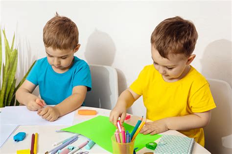 노란색과 파란색 셔츠를 입은 행복한 아이들이 책상에서 함께 예술과 공예품을 선합니다 아이들은 색깔의 종이에 그립니다 남자들은 집에서 놀고 있습니다 2 3 살에 대한 스톡