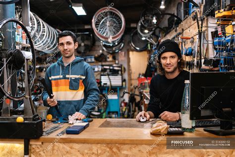 Positive Young Male Technician Assembling Bike While Colleague Working Positive Young Male Technician Assembling Bike While Colleague Working