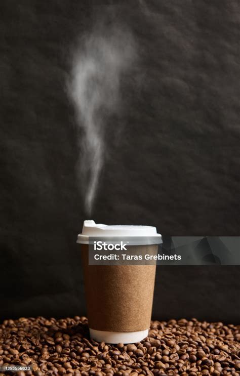 광고용 복사 공간이 있는 검은 벽 배경에 흩어져 있는 구운 커피 콩이 있는 표면에 증기가 있는 뜨거운 음료의 테이크아웃 골판지 컵의 클로즈업 구성 테이크아웃 음식에 대한