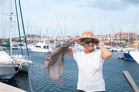 항구에서 선글라스를 착용하는 매력적인 시니어 여성 그는 도망치지 않도록 모자를 손으로 들고 있다 요트와 요트 그녀 주위 바람에 움직이는 스카프 경관에 대한 스톡 사진 및