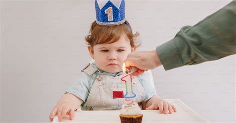 집에서 첫 생일을 축하하는 파란 왕관을 쓴 귀여운 사랑스러운 백인 소년 높은 의자에 앉아 컵케이크 디저트를 보고 있는 아이