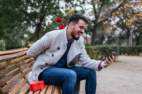 빨간 장미와 상자를 가진 행복한 남성은 공원 벤치에 앉아 스마트폰을 사용하고 있다 공원에 대한 스톡 사진 및 기타 이미지 공원 공원 벤치 긍정적인 감정 표현 Istock