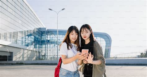 두 명의 예쁜 아시아 소녀가 공항 건물 앞에 서서 공동 비행 전에 스마트폰으로 메시지를 입력하거나 체크인을 하고 있다 사진 Unsplash의 여행 가방 이미지