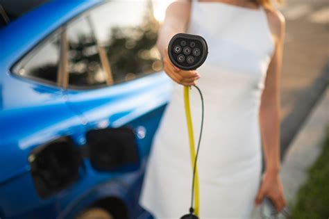 클로즈업 한 여자는 배경에 파란색 자동차와 함께 손에 전기 자동차 충전기를 들고 있습니다 30 34세에 대한 스톡 사진 및 기타 이미지 30 34세 교통수단 네트워크