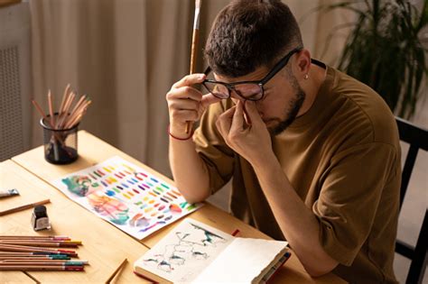 젊은 어두운 머리 피곤 한 남자의 초상화 안경에 수염을 가진 남성 예술가 카키 티셔츠 나무 테이블에 있는 스튜디오에 앉아 페인트 브러시를 손에 들고 앉아 있었다 피로로 눈을