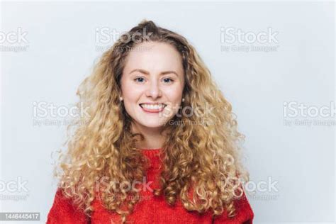 쾌적한 미소와 쾌활한 표정으로 행복한 곱슬 금발 여성의 실내 촬영 따뜻한 빨간 스웨터를 입고 흰색 배경에 대한 모델 사람 행복과 긍정적 인 감정 개념 곱슬 머리에 대한 스톡