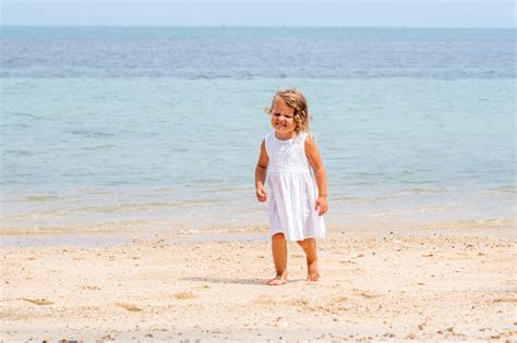 하얀 드레스를 입은 곱슬 머리를 가진 어린 소녀가 모래 사장을 걷습니다 한 아이가 해변을 따라 걷는다 아이들과 함께하는 여행 및 휴가 2 3 살에 대한 스톡 사진 및 기타