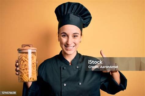 젊은 아름 다운 요리사 여자 입고 밥솥 유니폼 들고 항아리 와 이탈리아 파스타 마카로니 와 깜짝 얼굴 가리키는 손가락 에 자신 가리키기에 대한 스톡 사진 및 기타 이미지