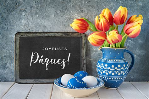 부활절 달걀과 회색 배경에 칠판이있는 파란색 세라믹 투수의 빨간색 노란색 튤립 텍스트 조이우스 Pýques는 프랑스어로 행복한 부활절을 의미합니다 0명에 대한 스톡 사진 및